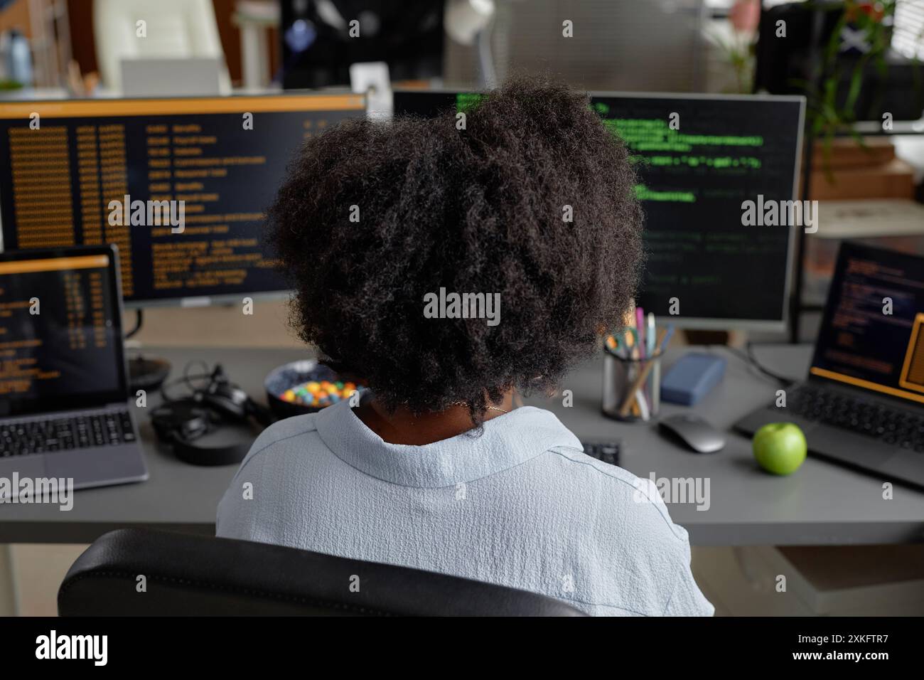 Rear view of female programmer of Black ethnicity working on multiple computers reading screen ...