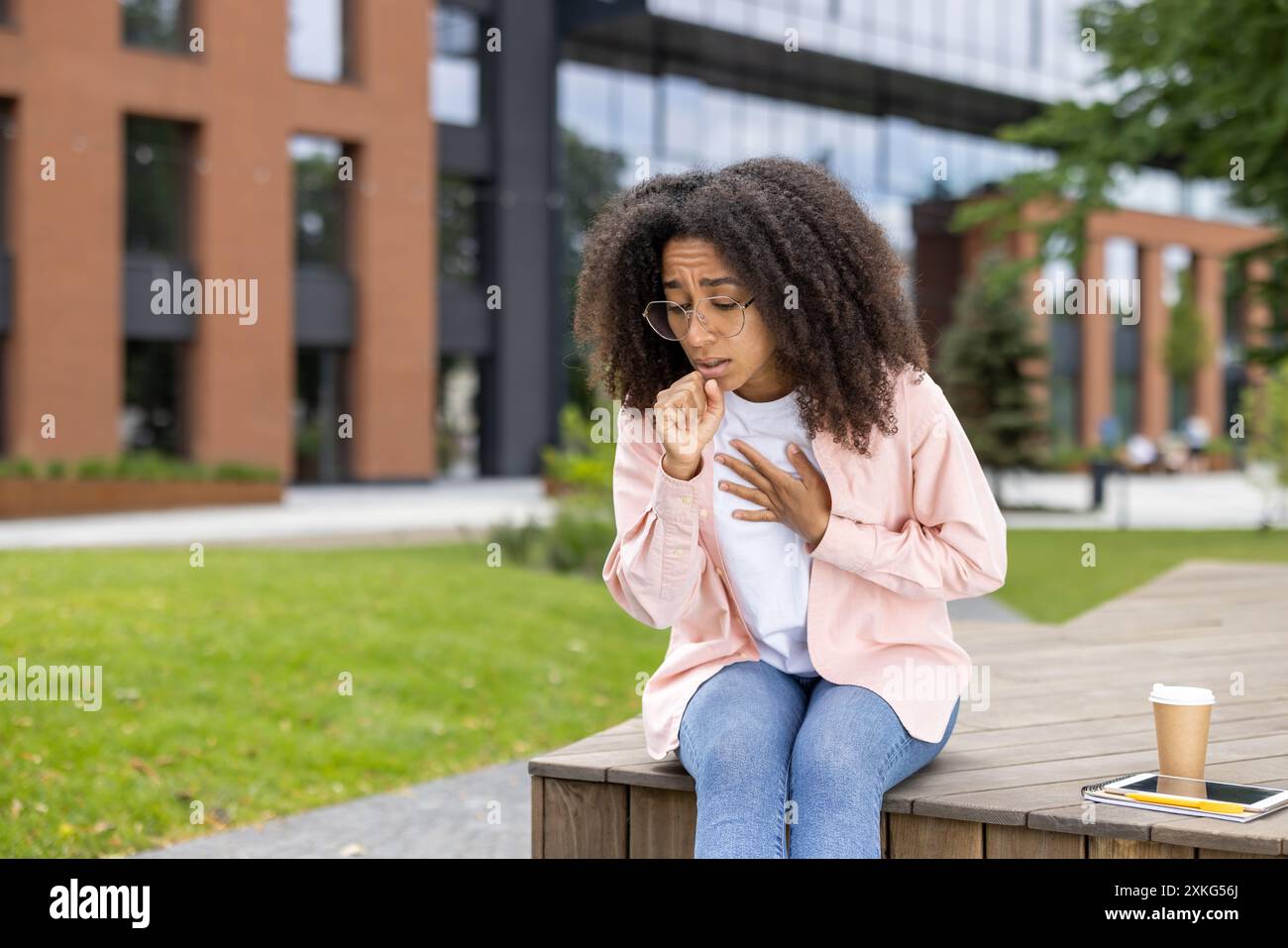 Young woman coughing while sitting outside urban building. Appears ...
