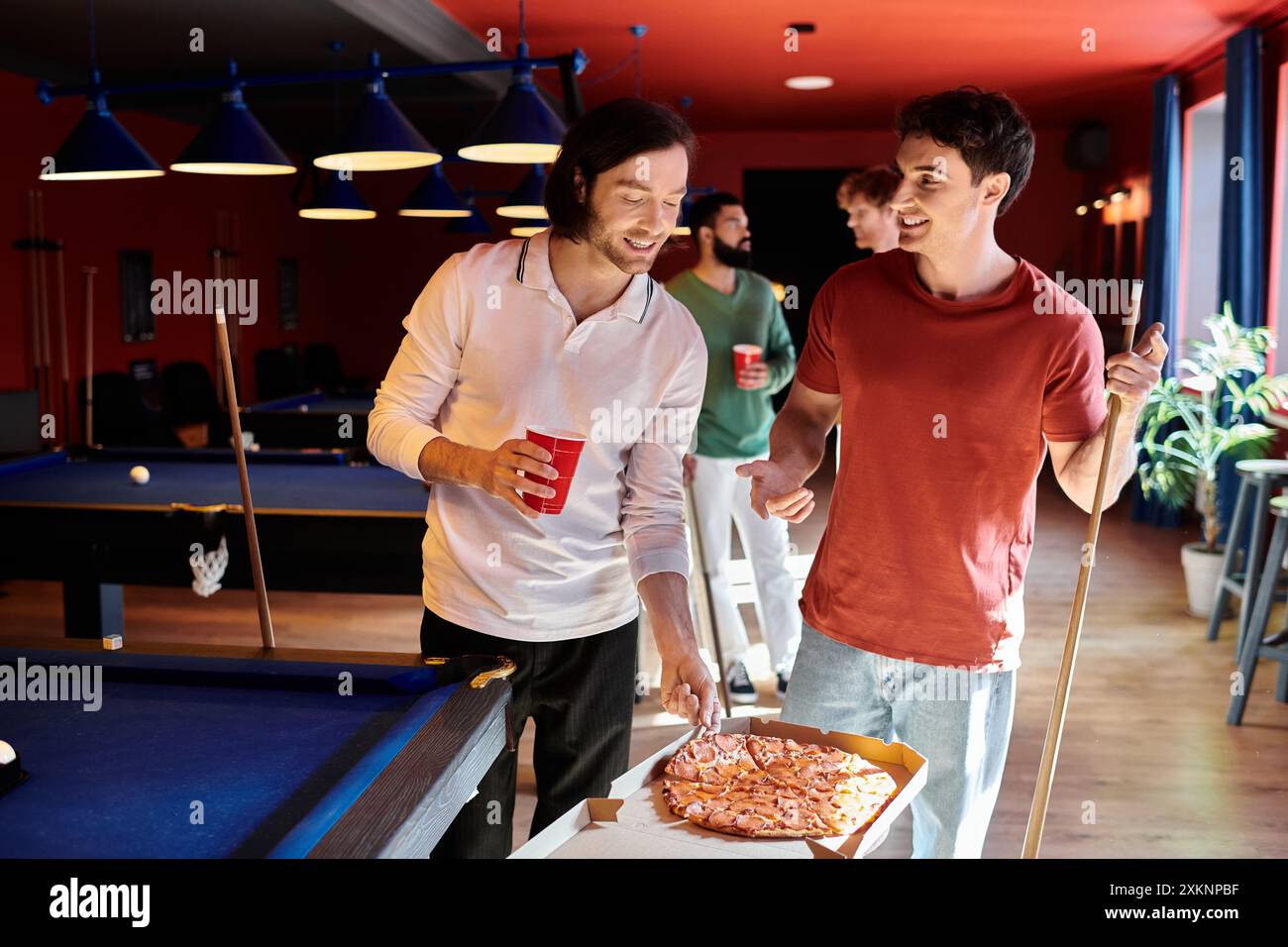 Friends stand by a pool table in a casual setting, enjoying pizza and ...