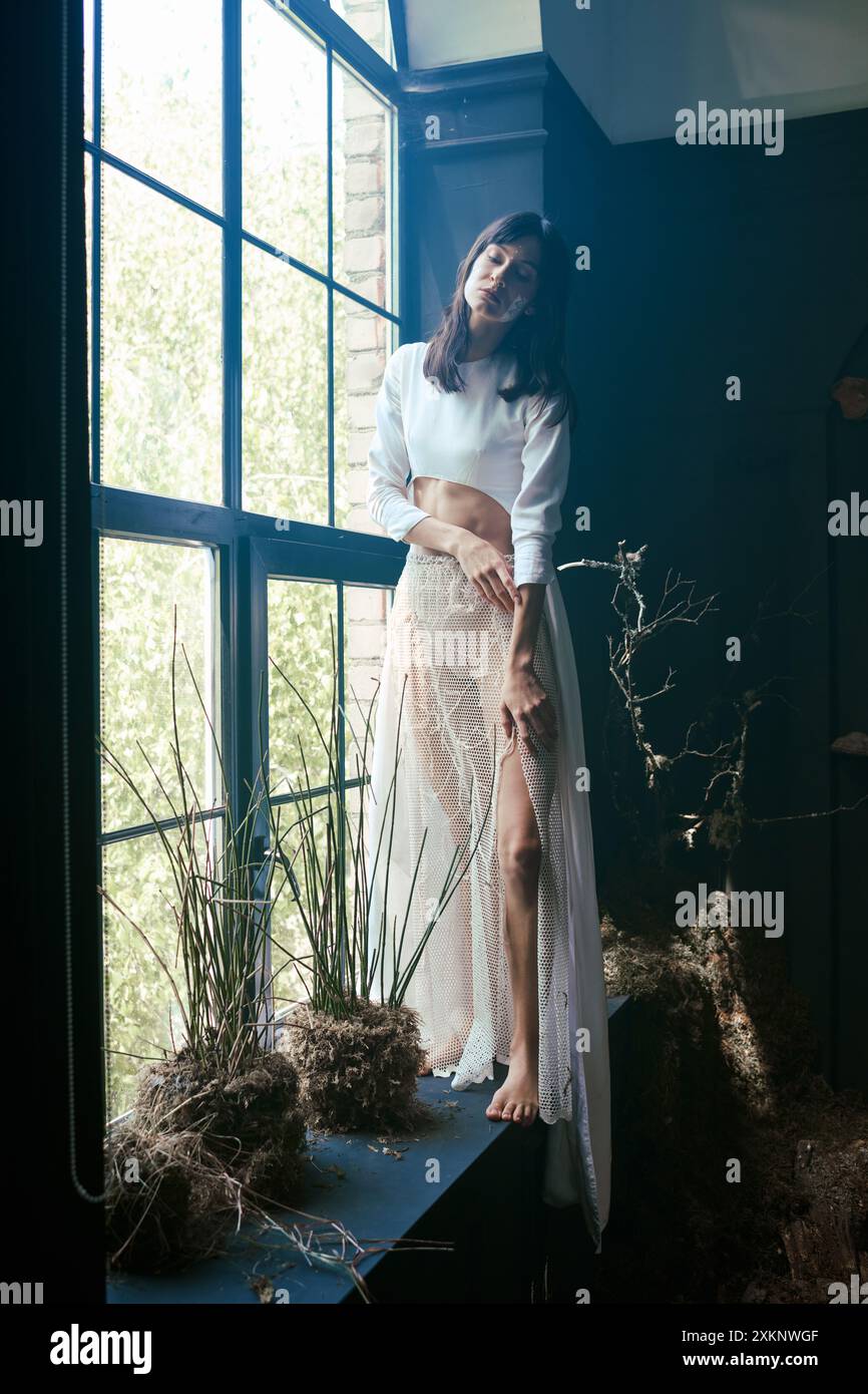 A woman in a white top and sheer skirt stands near a window with green ...