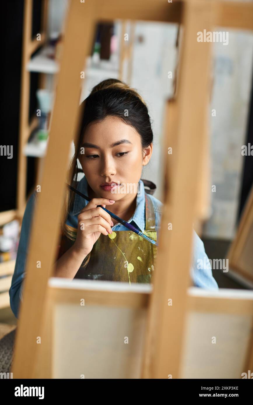 A young Asian artist, wearing an apron, thoughtfully paints in her ...