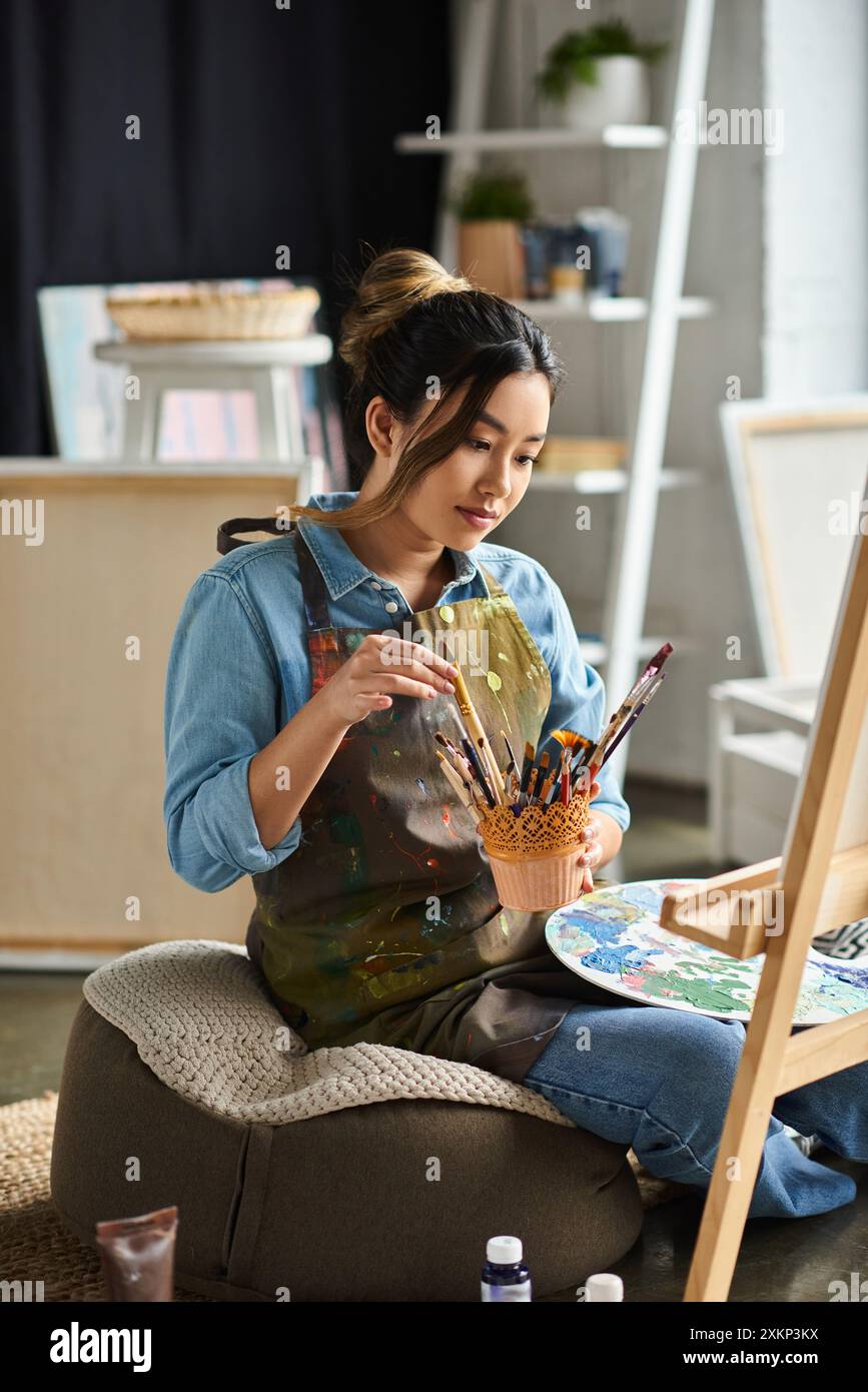 An Asian artist, wearing an apron, sits in her workshop and focuses on ...