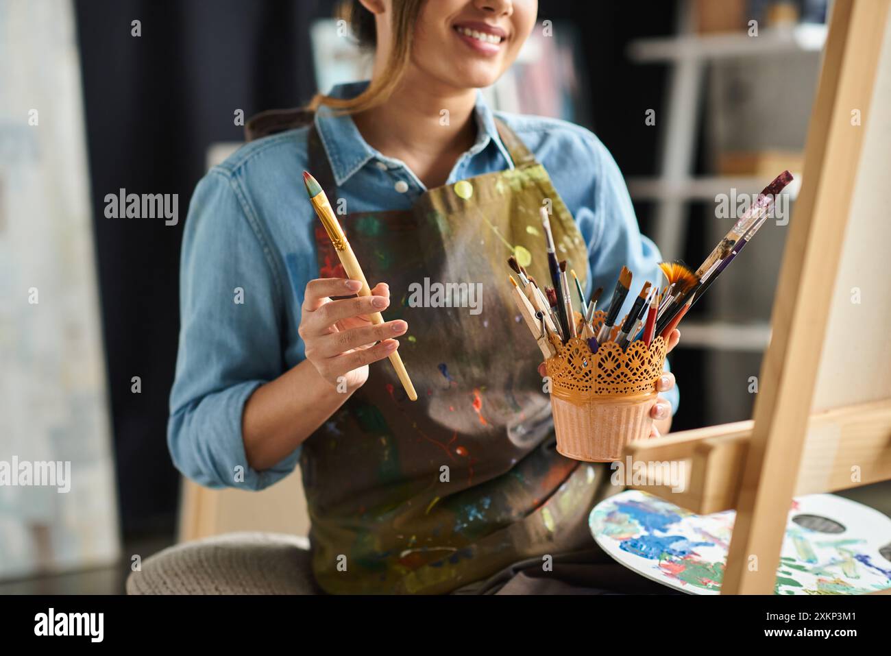A young Asian woman artist, wearing an apron, holds a paintbrush and ...