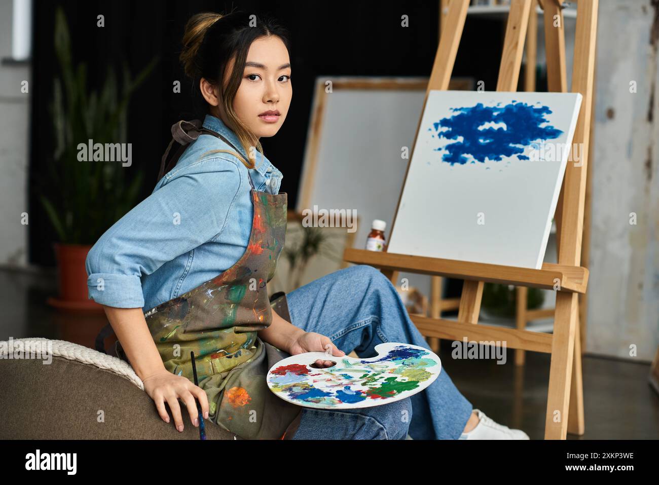 An Asian artist, dressed in a denim shirt and apron, sits in her ...