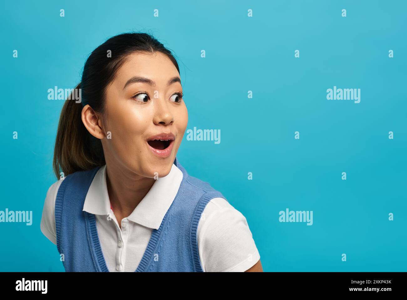 A surprised young Asian woman in casual attire stands against a blue ...