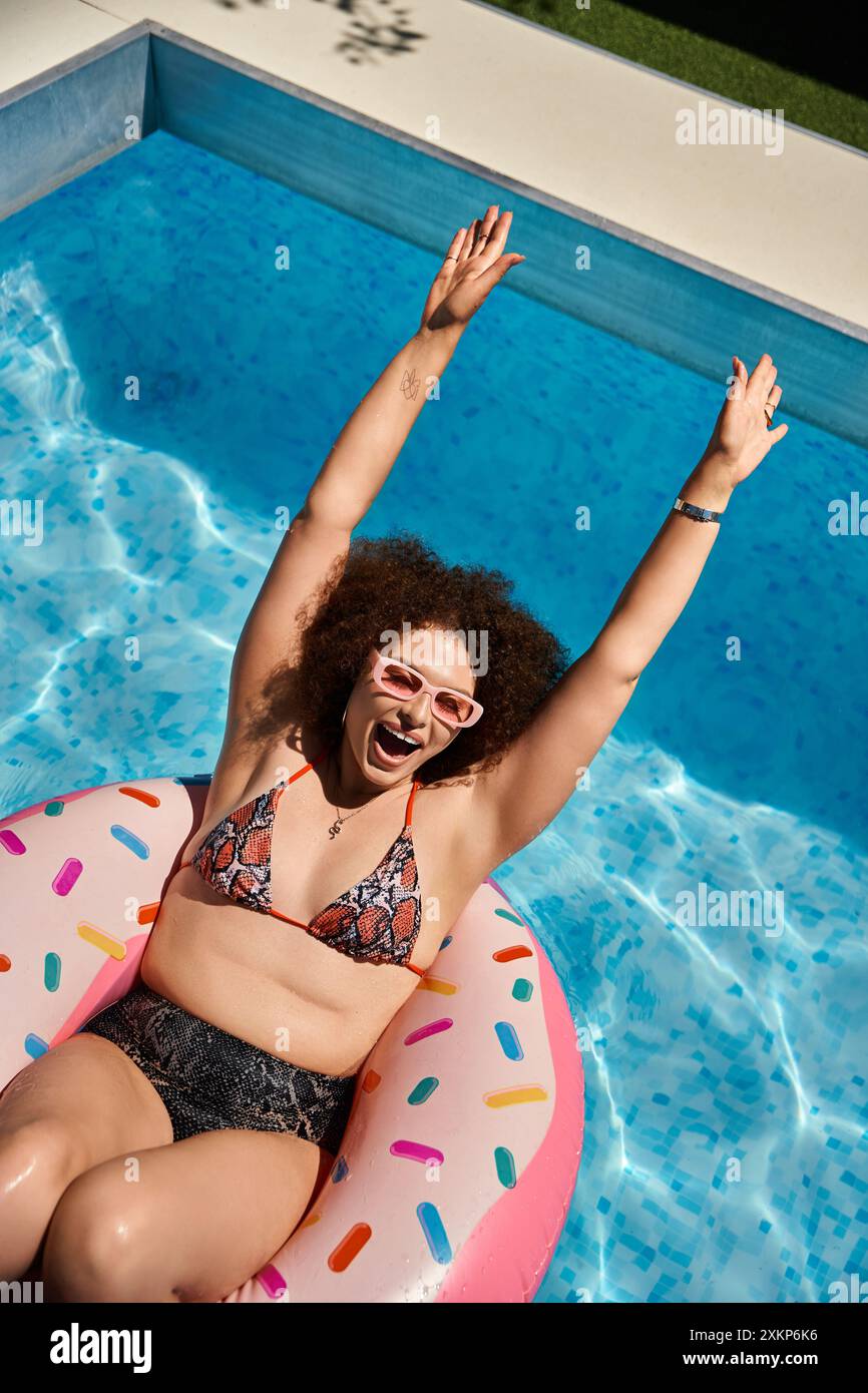 A woman with curly hair in a bikini floats on a donut-shaped pool float and smiles brightly ...