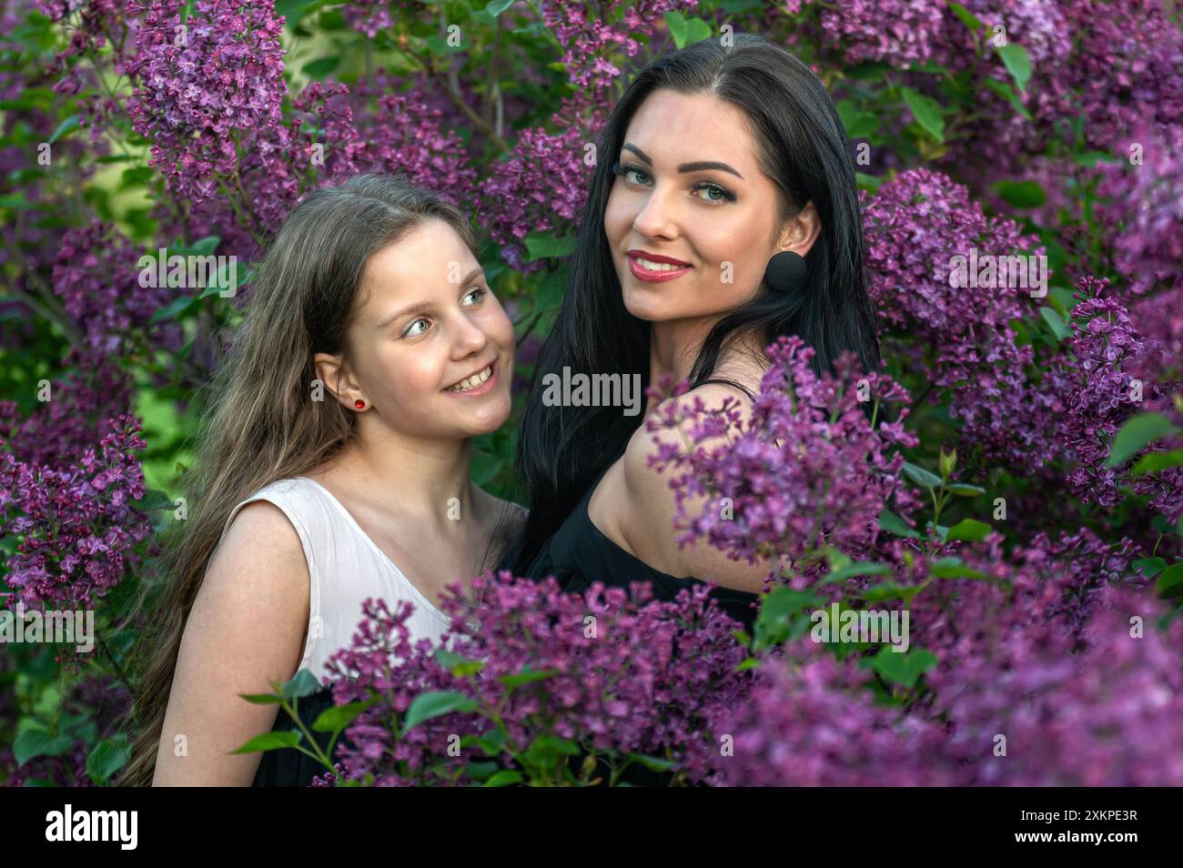 A woman and her sister smile and pose in front of blooming lilac bushes ...