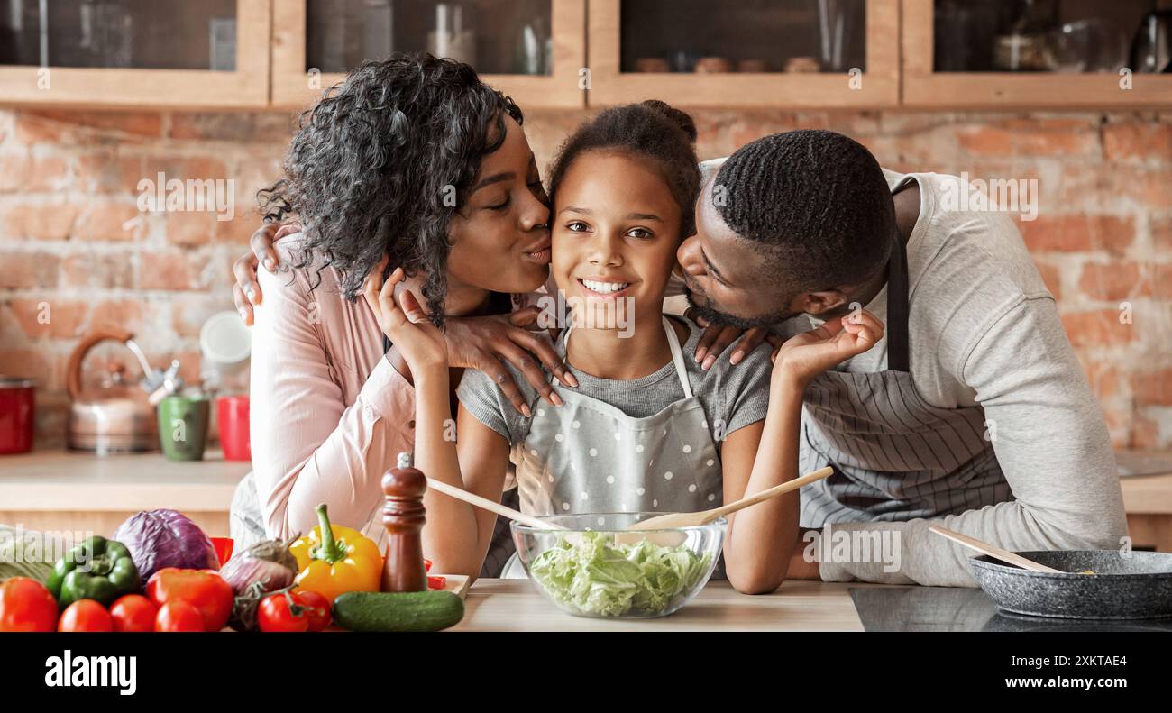 Adorable black family cooking healthy dinner together at kitchen, parents kissing daughter ...