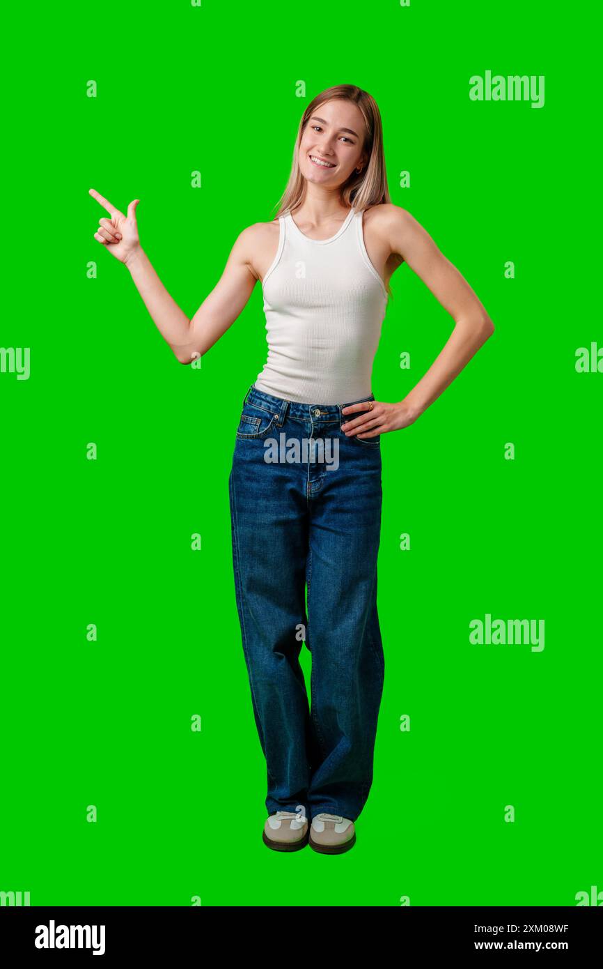 Woman in White Tank Top and Jeans Points to the Side Against a Green ...