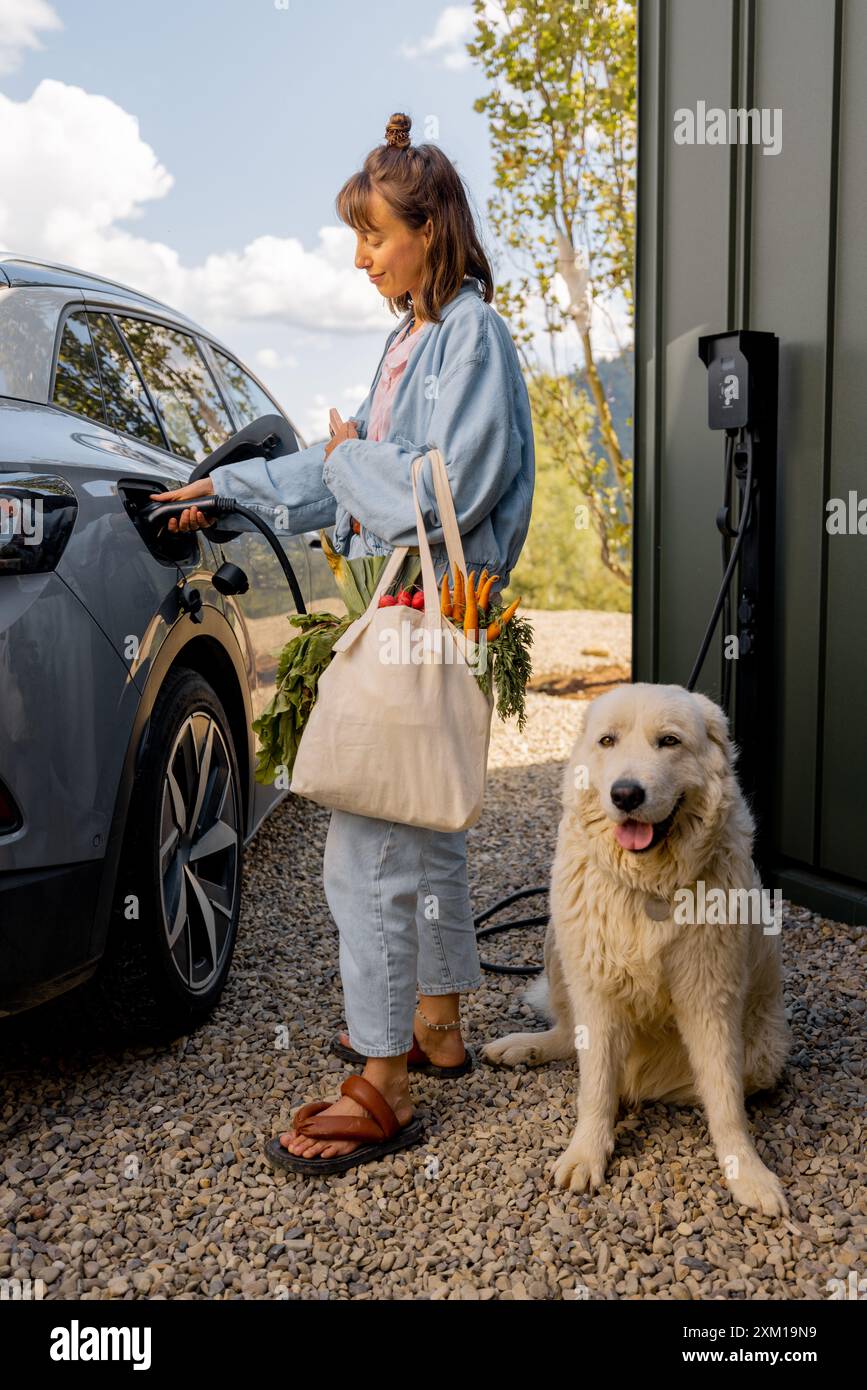 Woman plugging a charger into electric vehicle, with a dog near house ...