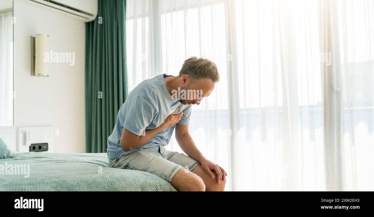 Middle-aged man sitting on bed, clutching chest in pain, heart attack ...