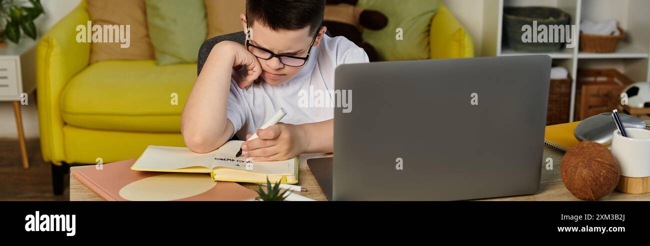Boy with Down syndrome writing in notebook at table, looking at laptop ...
