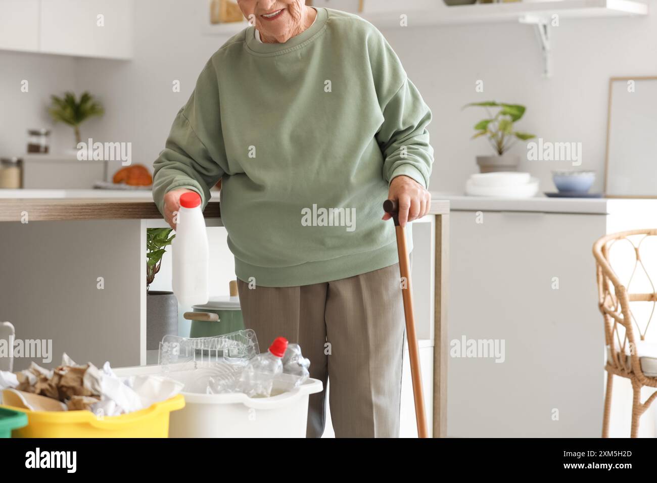 Senior woman throwing plastic bottle into garbage bin in kitchen. Waste ...