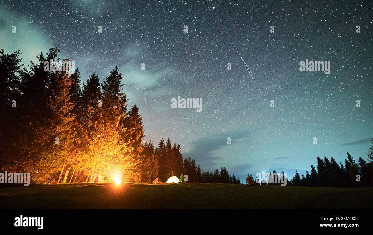 Campfire glows warmly beside several illuminated tents on grassy ...