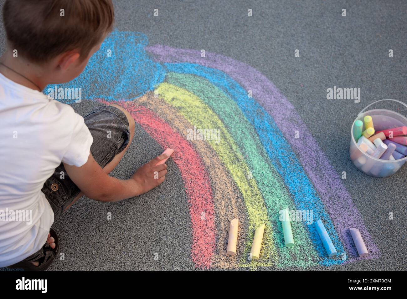 A boy draws a rainbow with multi-colored chalk on the asphalt. Drawing ...