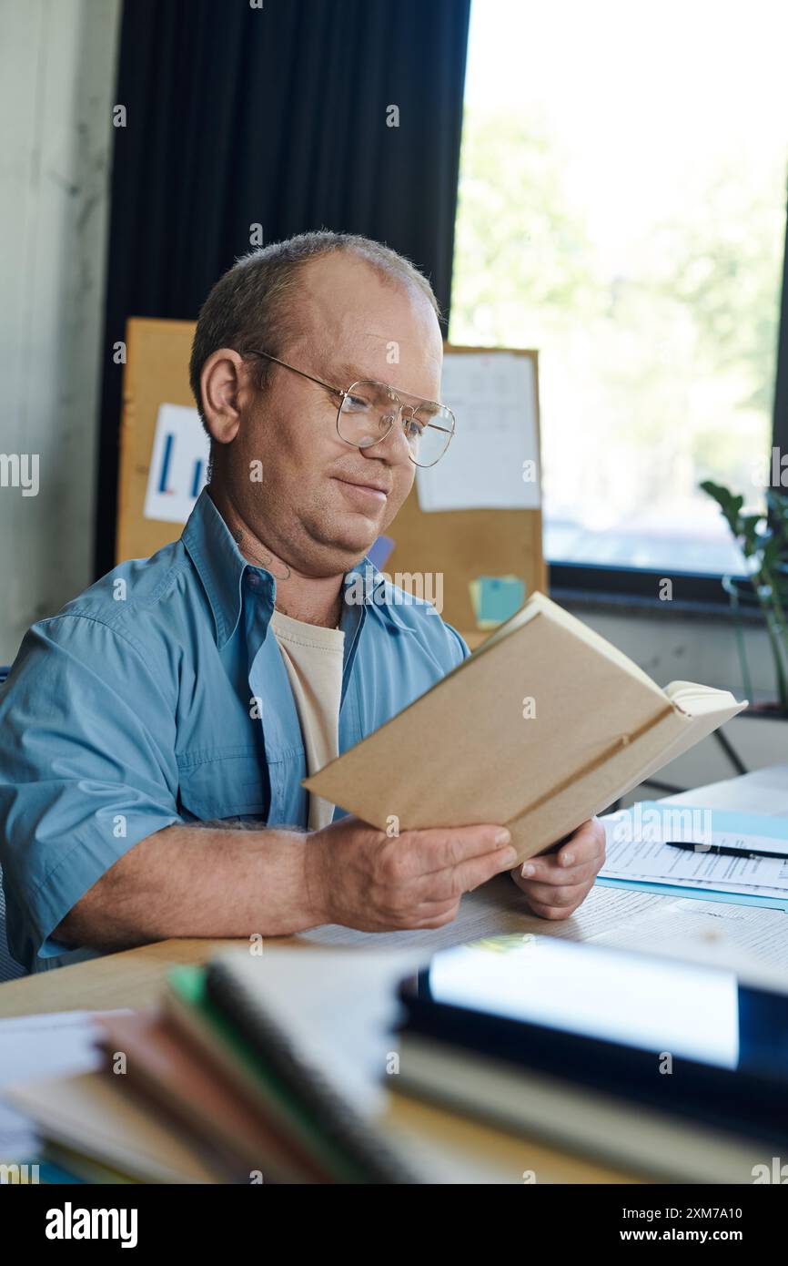 A man with inclusivity wearing glasses sits at a desk reading a book ...