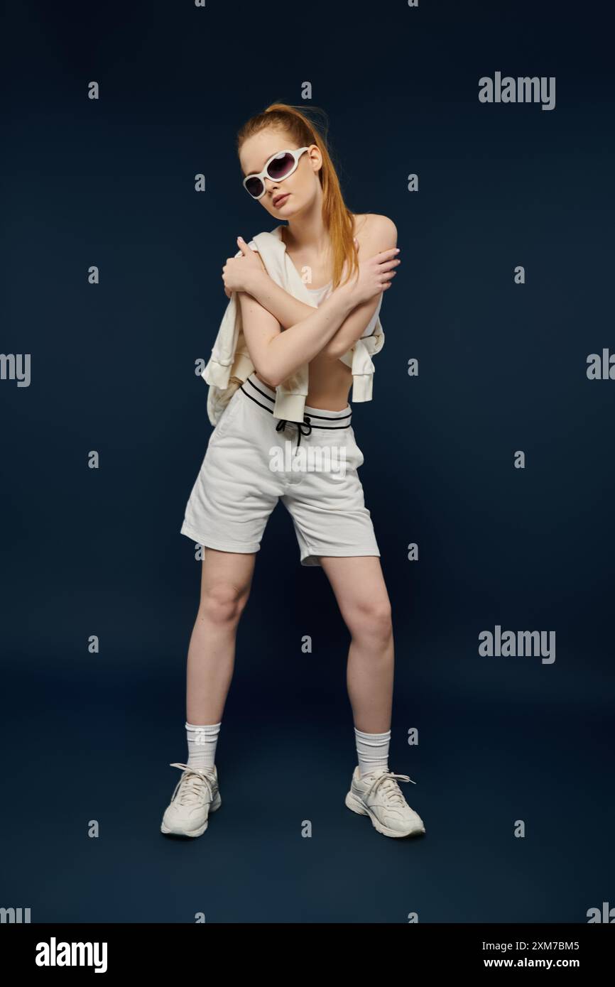 A young woman in white shorts and a white top poses against a dark blue ...