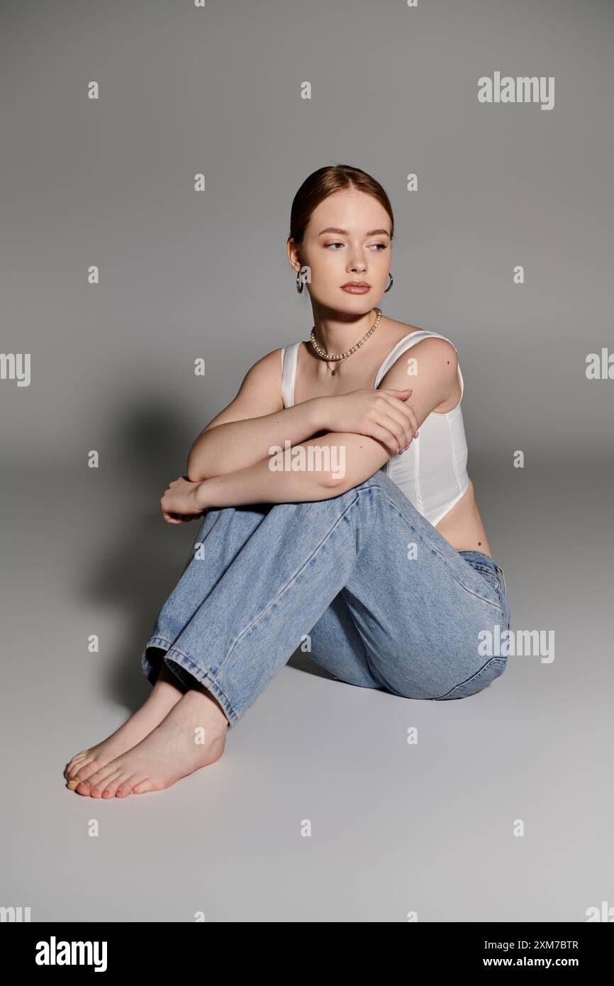 Woman in blue jeans and white top sits against grey backdrop, legs ...