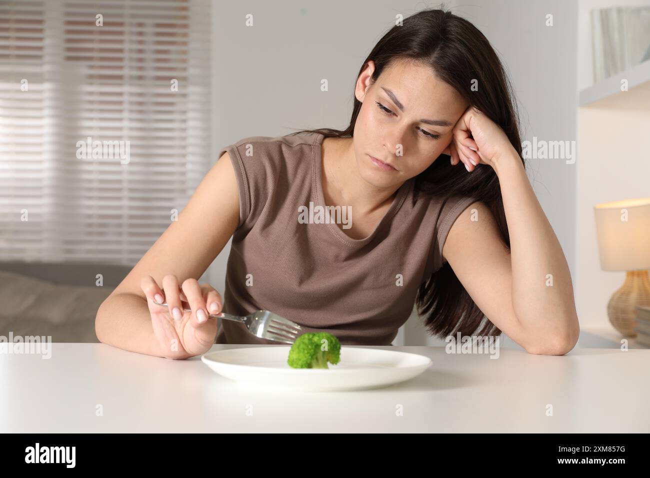 Eating disorder. Sad woman holding fork near broccoli at table indoors ...