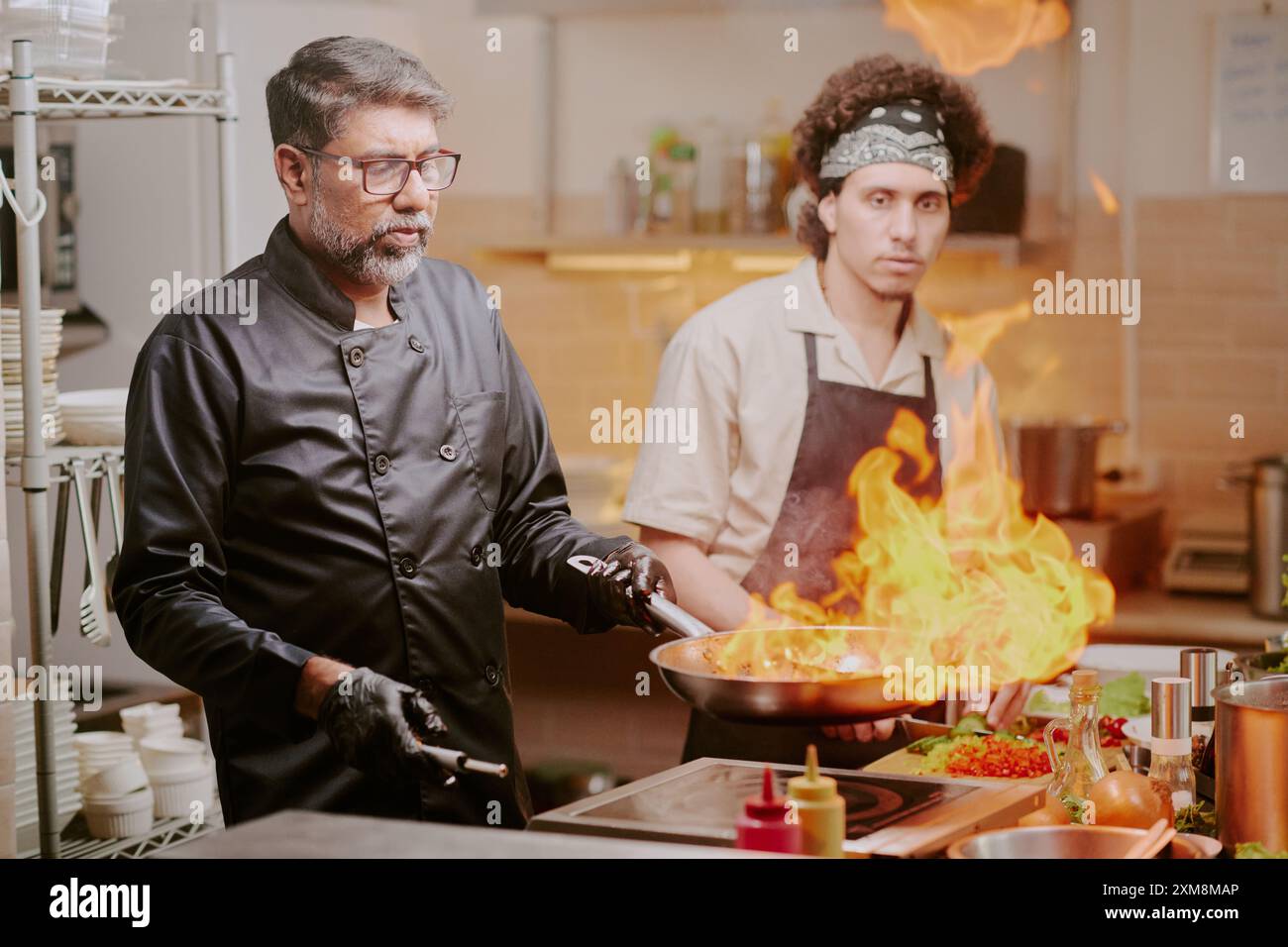 Middle aged chef cooking meat with fire while his best cook watching ...