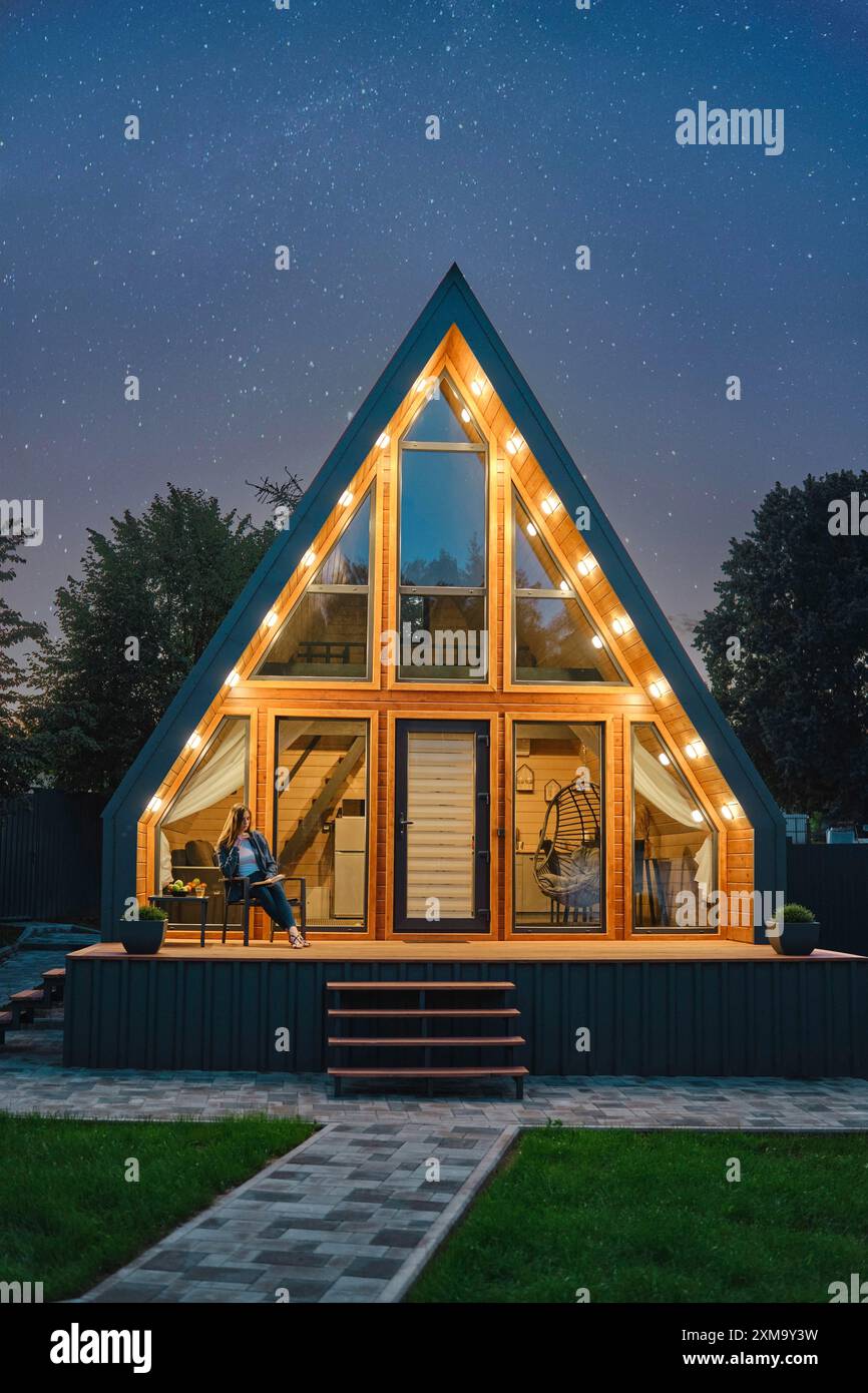A triangular log cabin illuminated from within stands under a starry ...