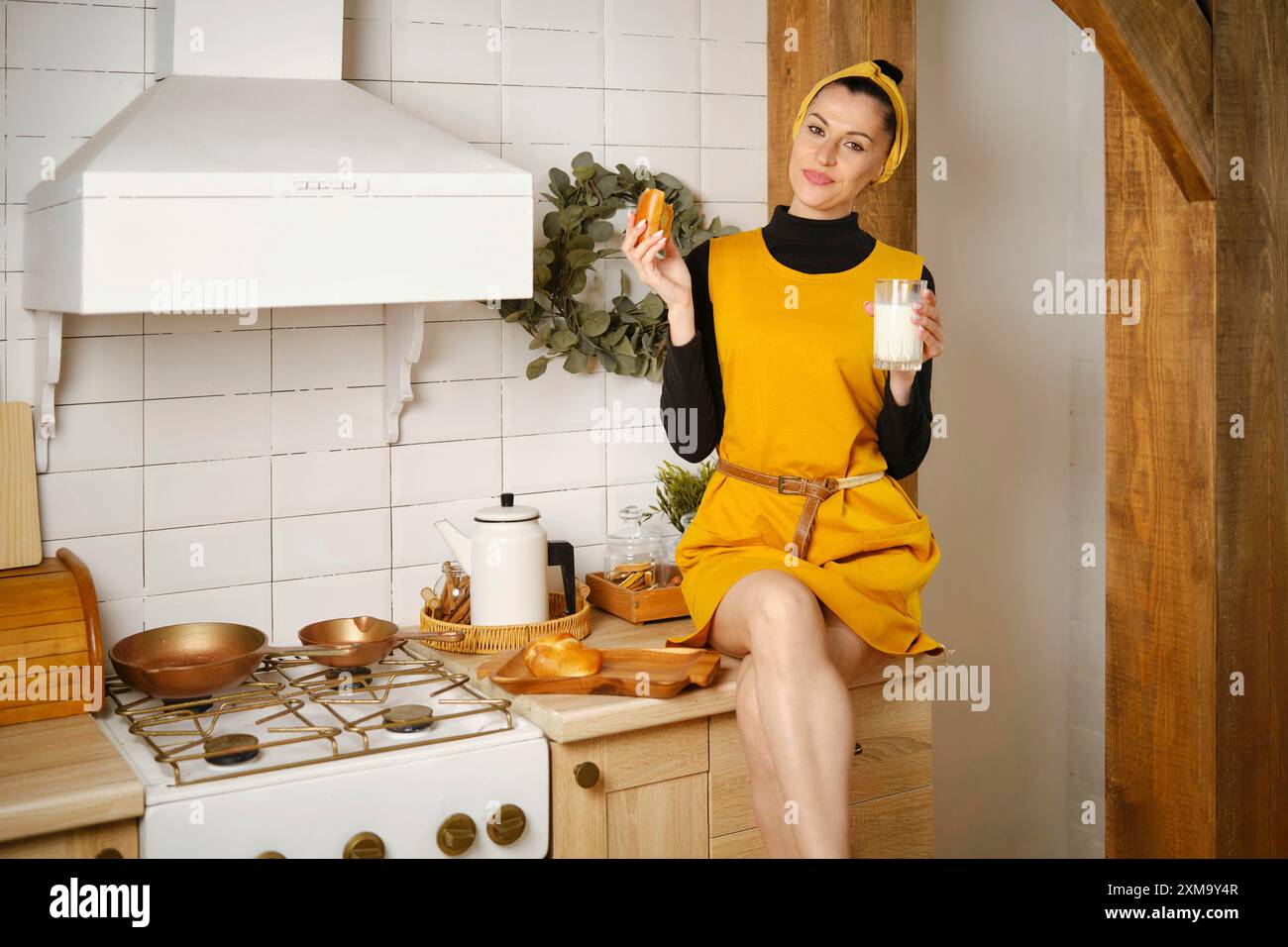 Cute woman sits in a kitchen drinks a glass of milk and eats bun. She ...