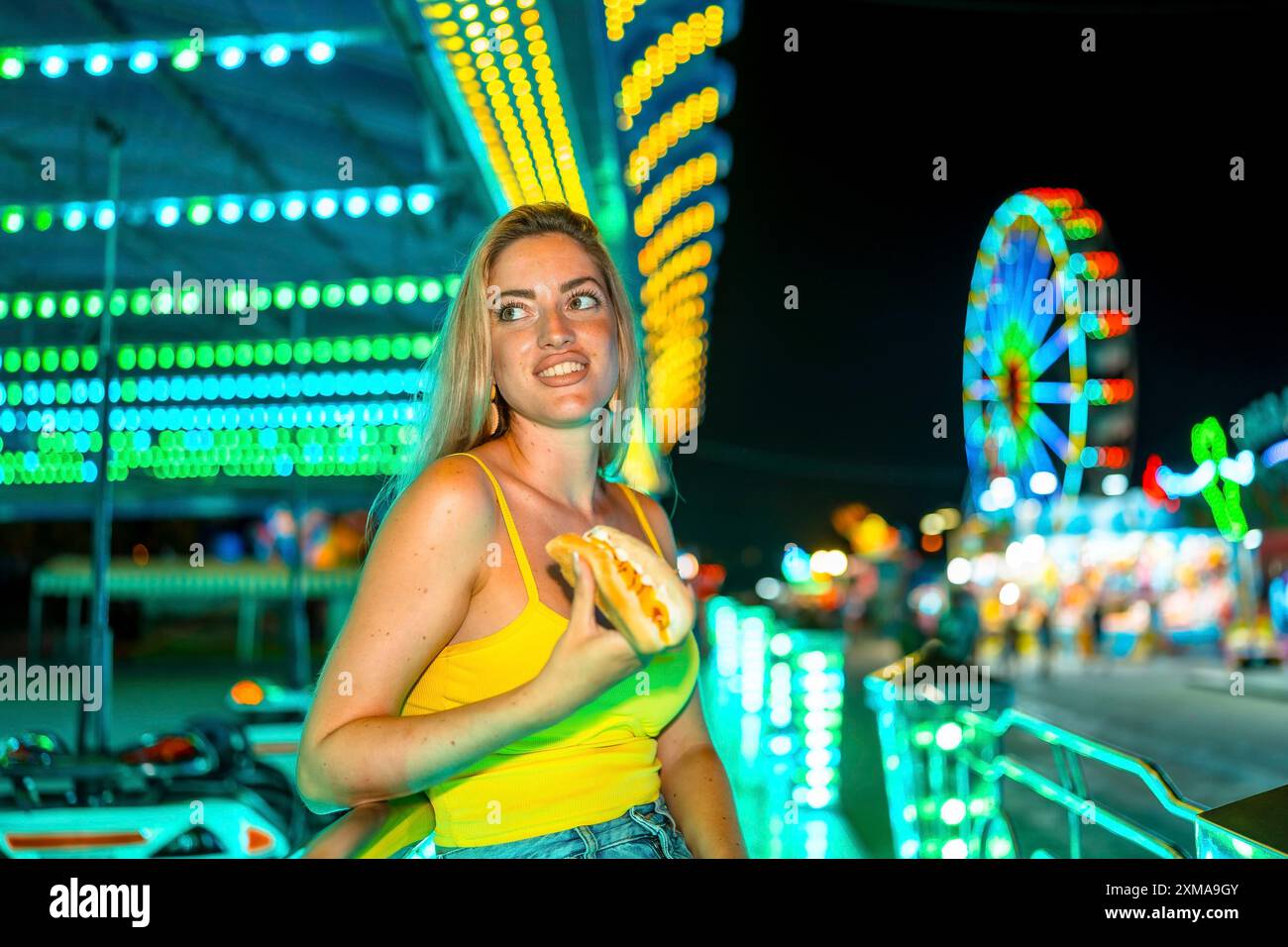 Blonde woman holding an unhealthy hot dog in a night fair in summer ...