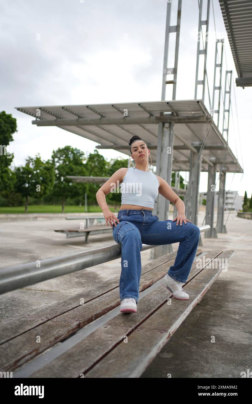 A woman is sitting on a bench in a park. She is wearing blue jeans and ...