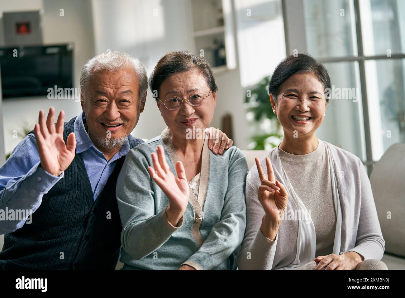 group of three happy senior asian people sitting on couch at home waving hands to camera while ...