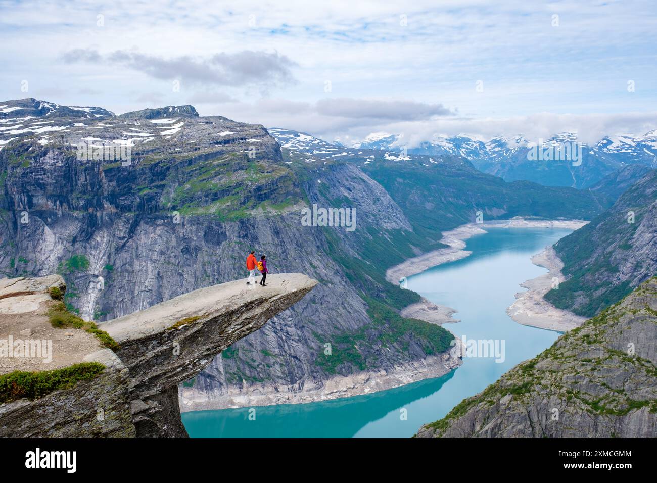 Two hikers stand on a cliff edge overlooking a dramatic fjord and ...