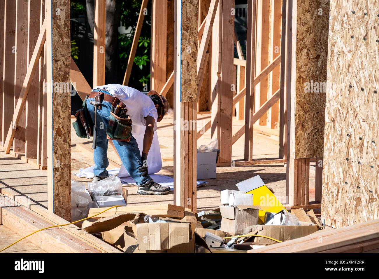 Construction worker in toolbelt and hard hat reviewing construction drawings on new house wood ...