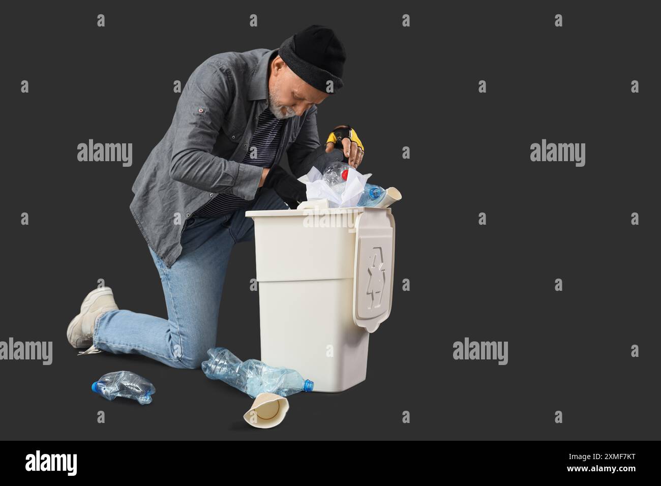 Mature homeless man rummaging through garbage bin on dark background ...