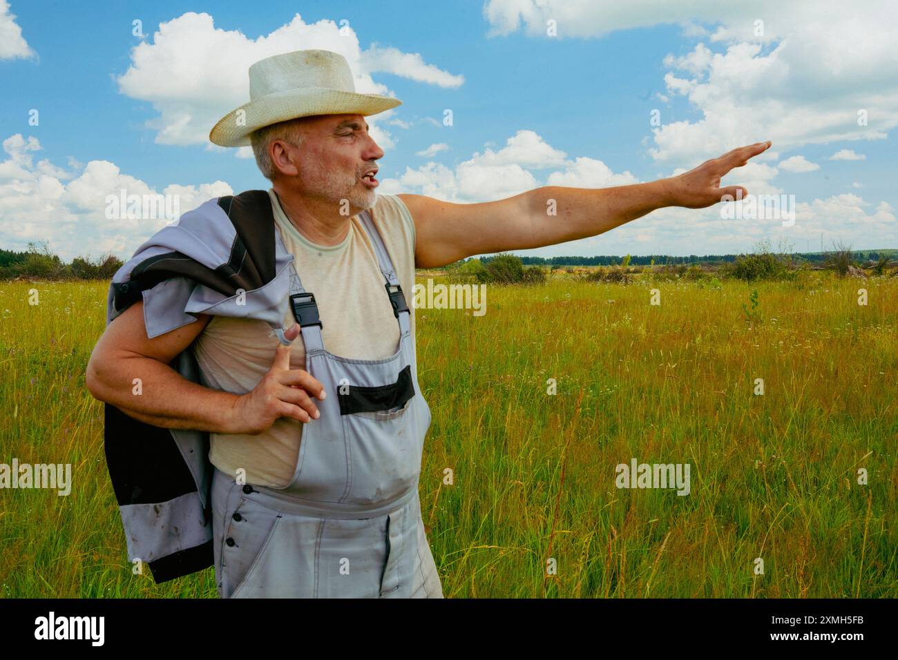 An elderly farmer in a hat stands in a grassy field, pointing towards ...
