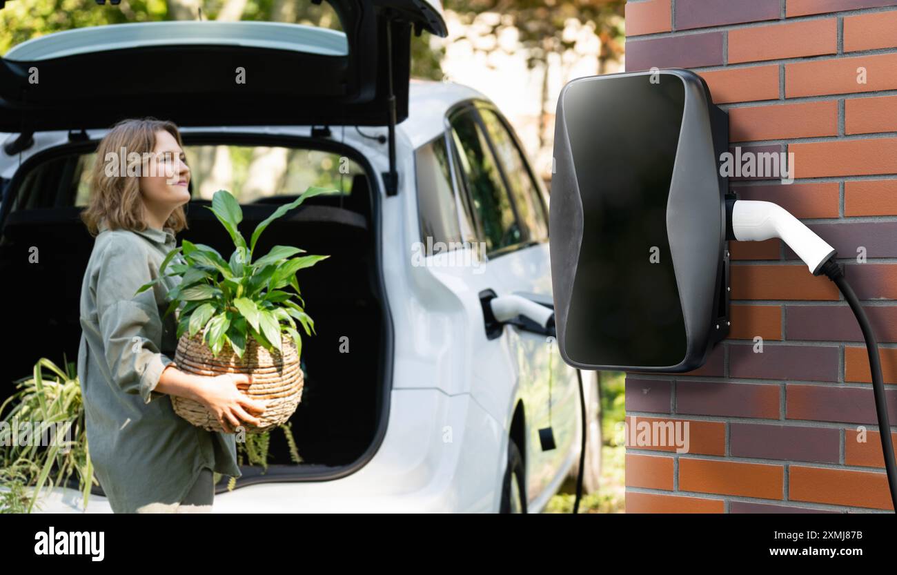 Woman with plant in pot next to a charging electric car in the yard of ...