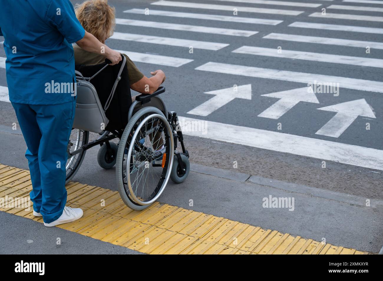 Rear view of a nurse helping an elderly woman in a wheelchair cross the ...