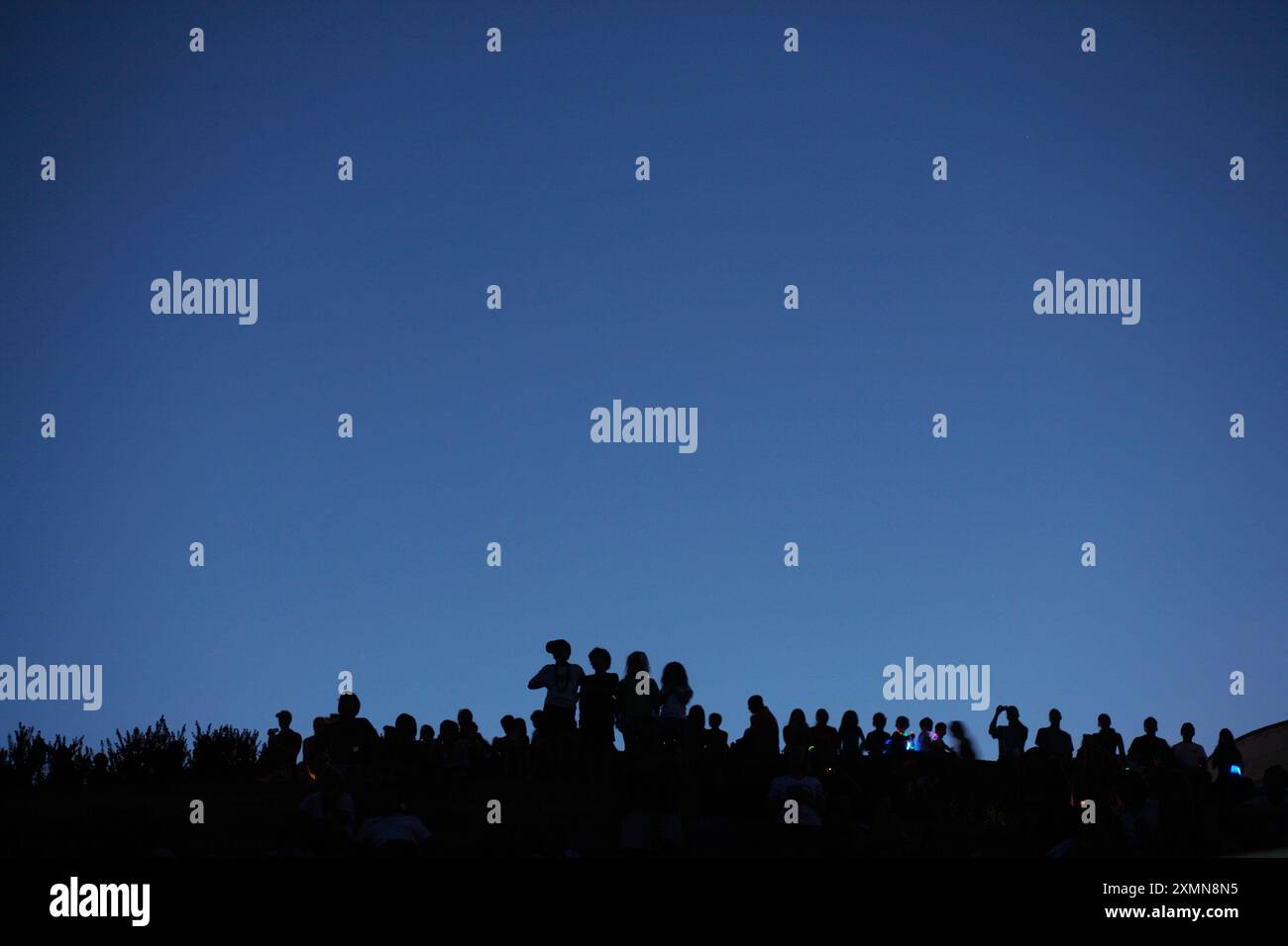 A crowd of people are silhouetted against a blue evening sky as they ...
