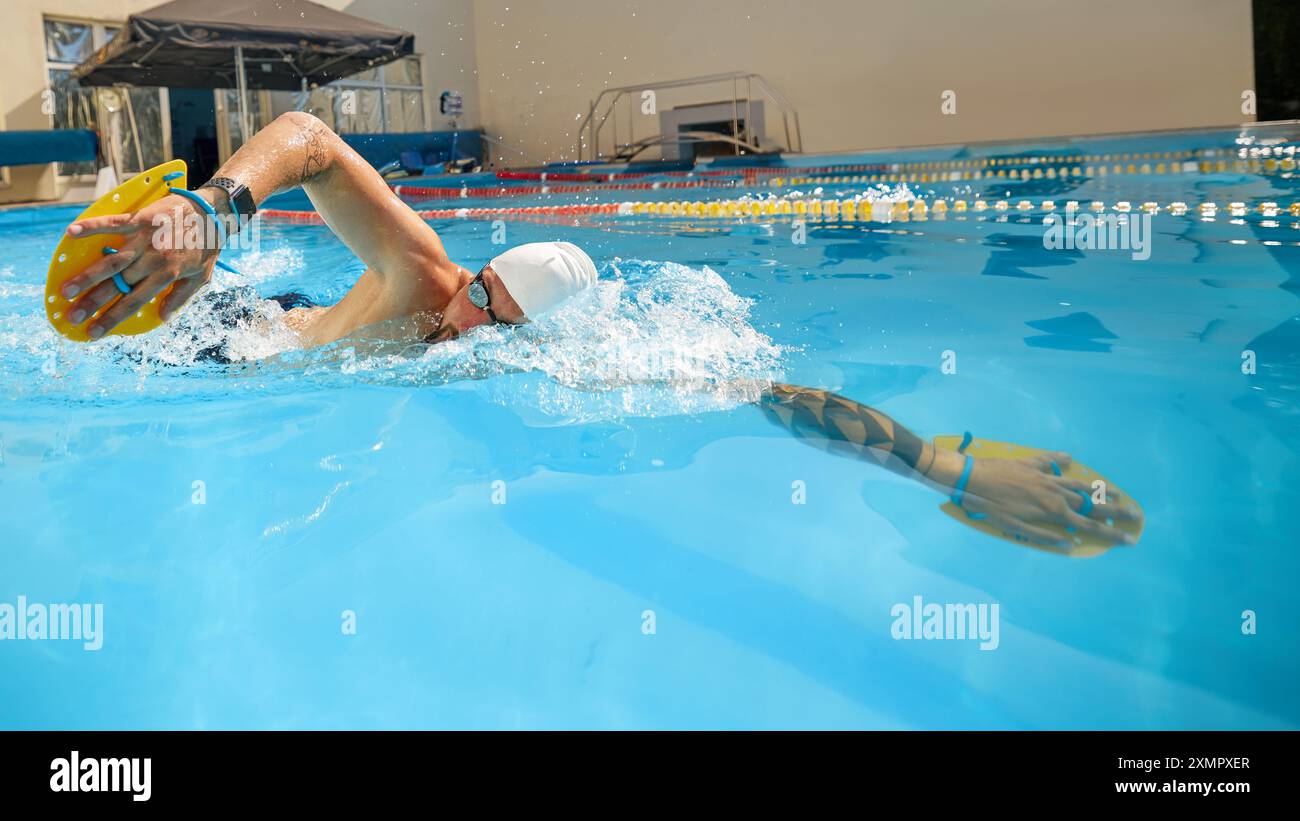 Swimmer wearing white cap, and yellow paddles trains in clear blue pool ...