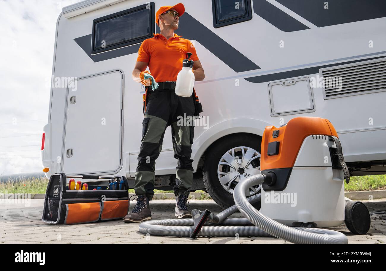 A worker in an orange uniform stands by a camper van, holding cleaning ...