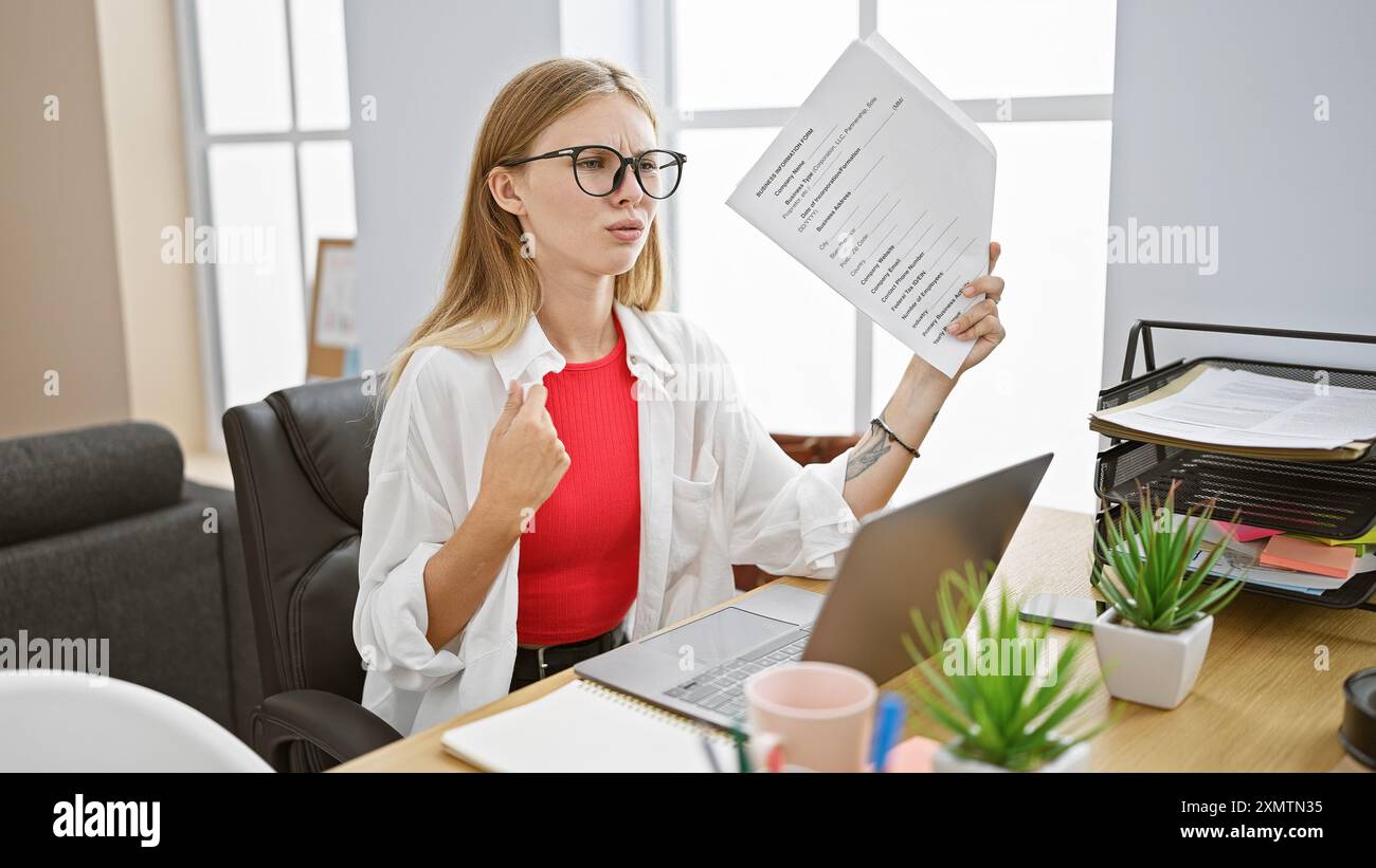 Blonde woman in office fanning herself with paper, looking tired ...