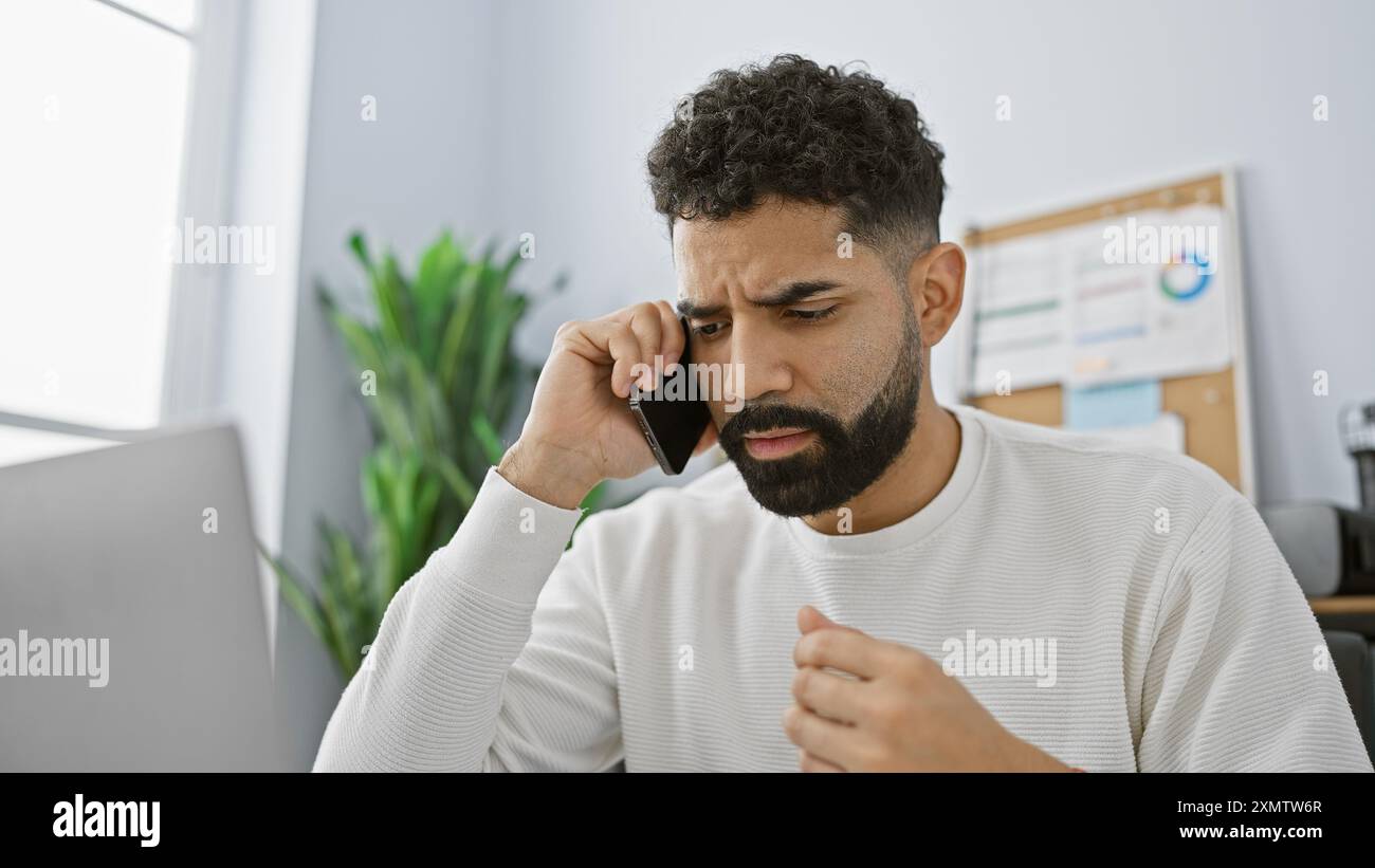 A bearded man talks on the phone in an office, showing concern while ...