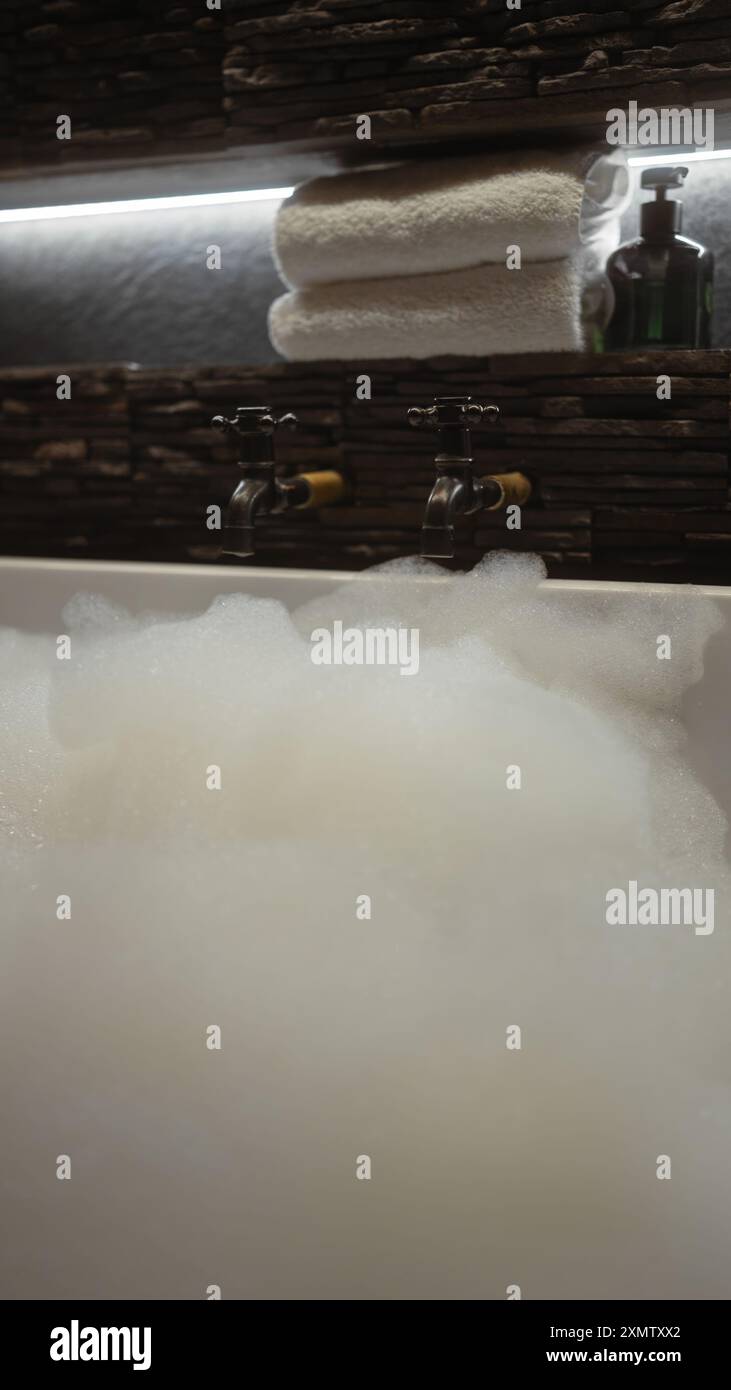 Overflowing bathtub under lit mirror shelf with towels and soap bottle ...