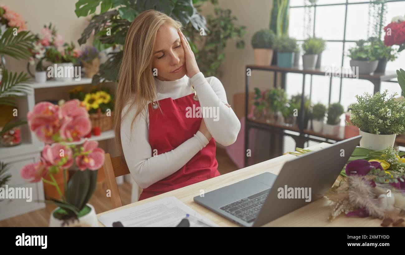 Young caucasian woman in apron feeling tired at flower shop with laptop ...