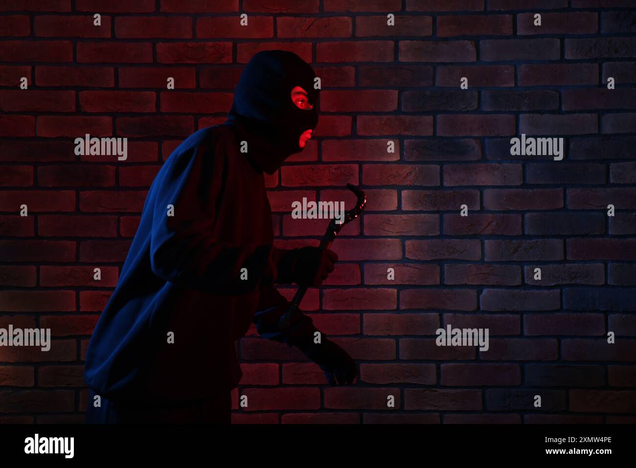 Thief in balaclava with crowbar in color lights against red brick wall ...