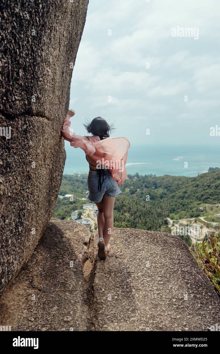 A woman walks along a narrow cliff edge, her back to the camera, with ...