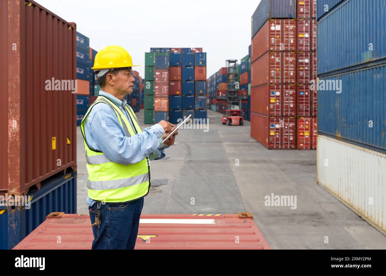 A worker inspects shipping containers in a busy port, wearing a safety ...