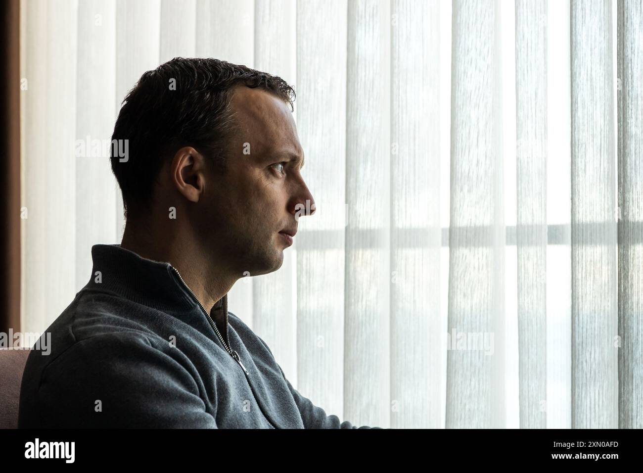 Serious young adult Caucasian man sitting in front of bright window ...