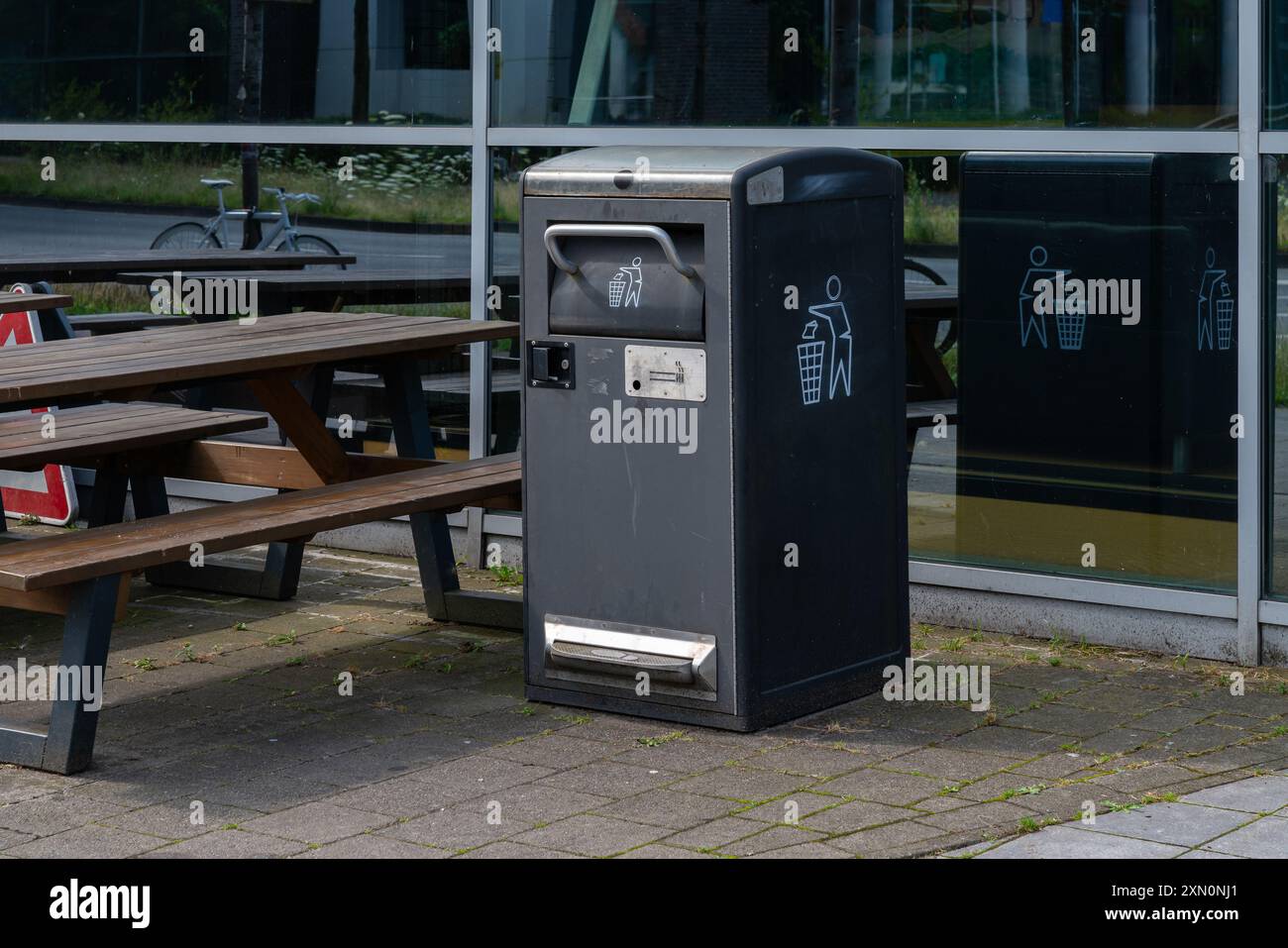 A black trash bin stands beside a wooden picnic table in an urban area ...