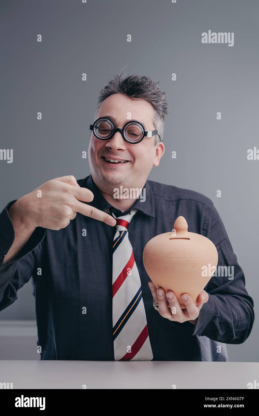 Businessman wearing thick funny glasses is pointing at a piggy bank he ...