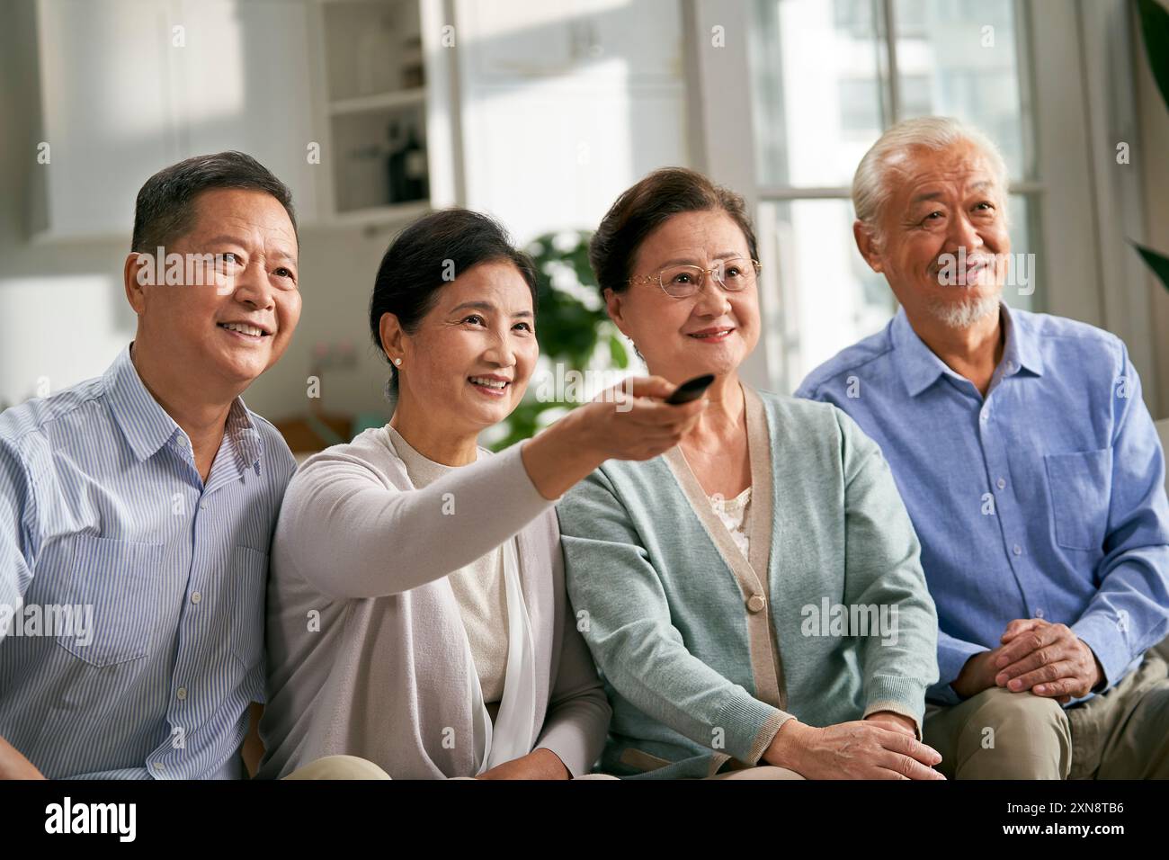 group of happy senior asian people two couples sitting on couch at home watching tv together ...
