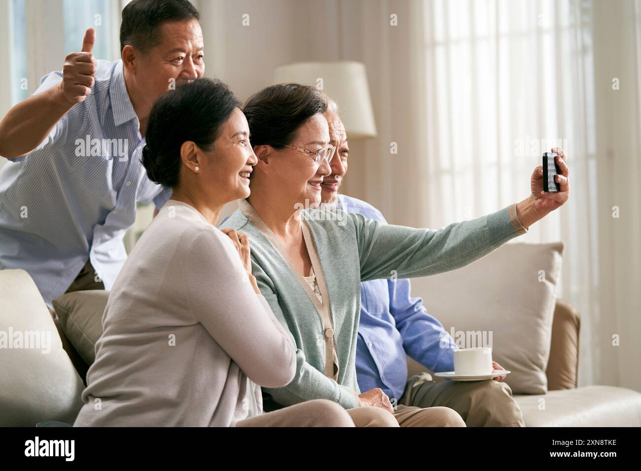 group of happy senior asian people two couples sitting on family couch at home taking a selfie ...